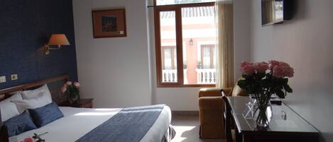 Quarto de hotel com cama, mesa de cabeceira, escrivaninha, cadeira e janela com vista para edifícios.