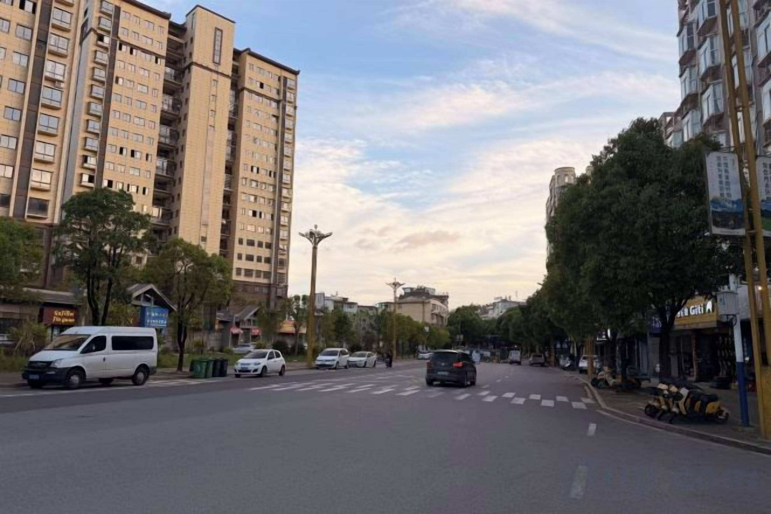 Um edifício com vários andares e fachada plana, com uma rua movimentada e árvores ao longo da calçada.