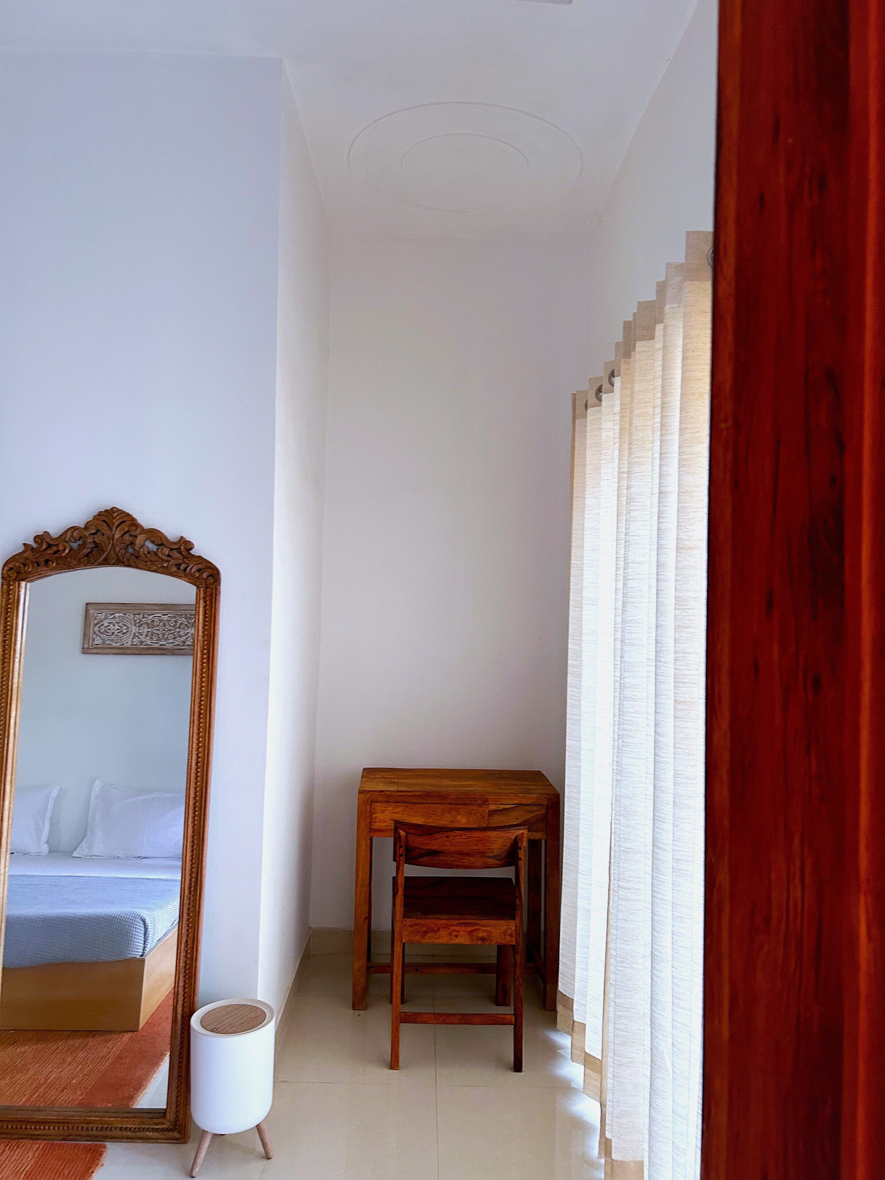 Quarto com cadeira de madeira, uma mesa pequena, um espelho e uma cama com roupa de cama branca.