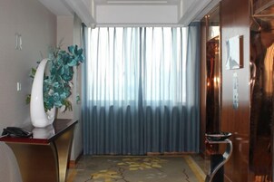 Um saguão de hotel com balcão de recepção, uma planta, uma janela grande com cortinas e uma parede de madeira.