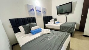 Quarto de hotel com duas camas, cada uma com cabeceira escura e roupa de cama cinzenta. Há uma TV de tela plana fixada na parede acima das camas.