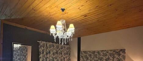 Quarto com teto de madeira, uma cama grande com colcha azul escura, duas luminárias de cabeceira, um lustre, um espelho e cortinas com padrão floral.