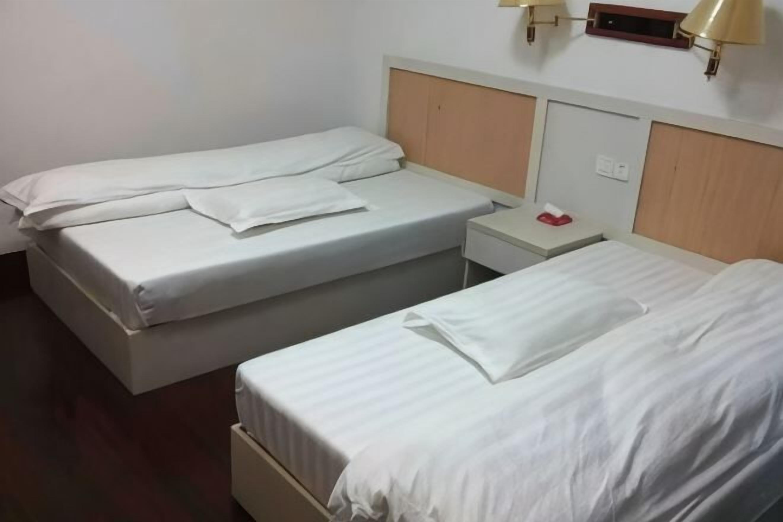 Duas camas de solteiro com roupa de cama branca, uma cabeceira de madeira, uma mesa de cabeceira com uma luminária e um objeto vermelho sobre ela.