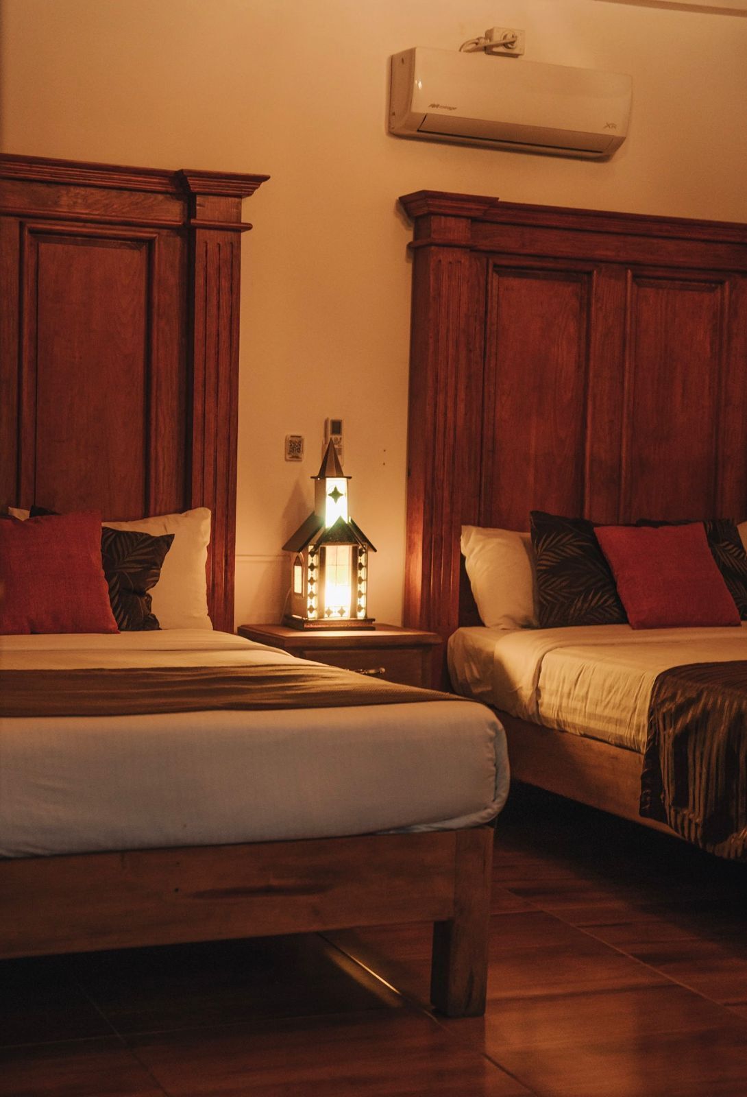 Quarto com duas camas de madeira, uma mesinha de cabeceira com um candeeiro e um aparelho de ar condicionado na parede.