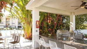Uma área de bar ao ar livre com churrasqueira, assentos e plantas tropicais.