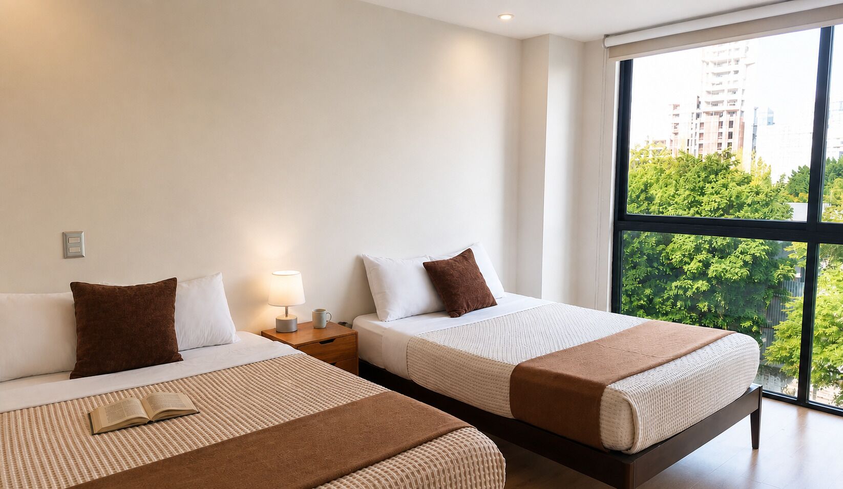 Quarto de hotel com duas camas, mesa de cabeceira com abajur e uma grande janela com vista para edifícios e árvores.