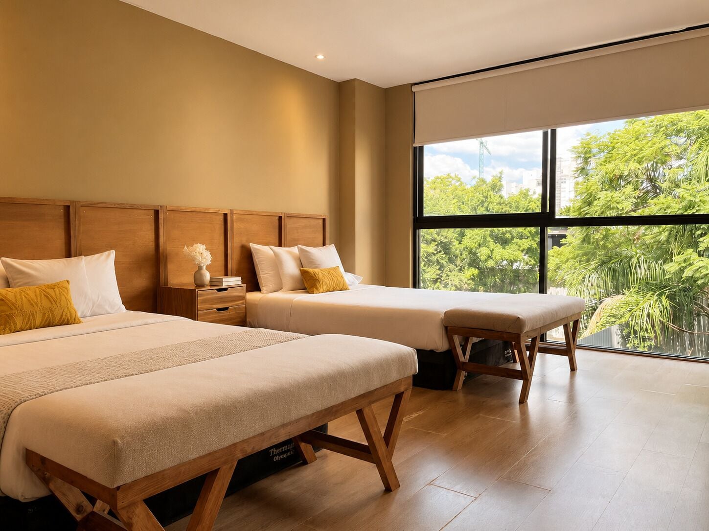 Quarto de hotel com duas camas, cabeceira de madeira, mesa de cabeceira e uma janela grande com vista para o verde.