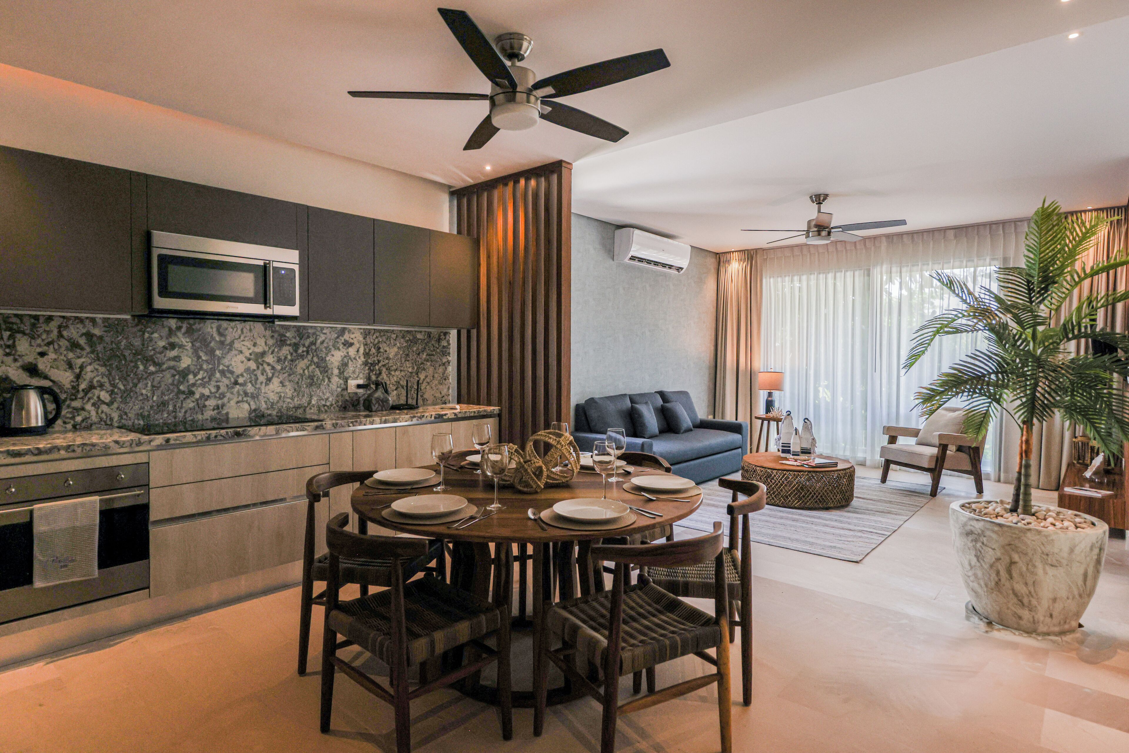 Cozinha moderna com micro-ondas, forno e área de jantar com cadeiras e mesa posta para quatro pessoas. Há uma sala de estar com sofá, mesa de centro e uma grande planta em vaso.