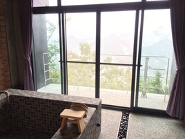 Casa de banho com banheira, um pequeno banquinho de madeira e vista para uma varanda com grades e vegetação.