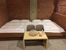 Uma cama de solteiro com roupa de cama branca, uma pequena mesa de madeira com artigos de higiene pessoal e uma parede de tijolos.