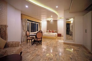 Quarto de hotel moderno com área de jantar, uma cama grande e banheiro com porta de vidro.