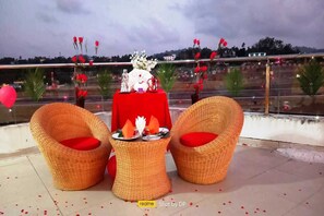 Uma área de refeições com cadeiras de vime e uma mesa posta para dois, com toalha de mesa vermelha e um adorno decorativo branco.