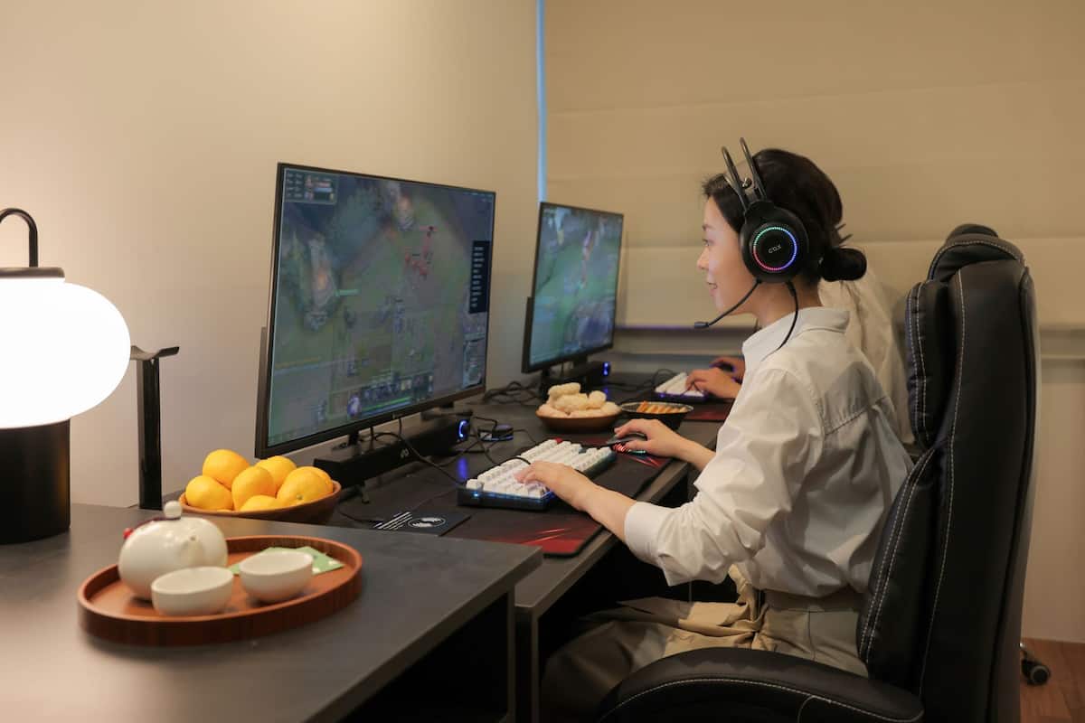 Uma pessoa sentada em uma mesa com dois monitores, um teclado e um fone de ouvido. Há uma tigela de laranjas e uma bandeja com bules na mesa.