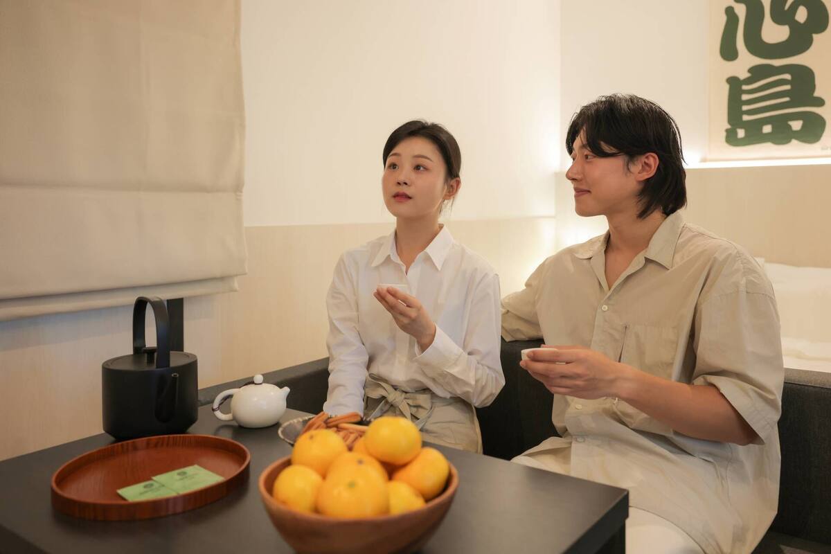 Duas pessoas sentadas em um quarto com uma mesa que tem uma tigela de frutas e uma chaleira.