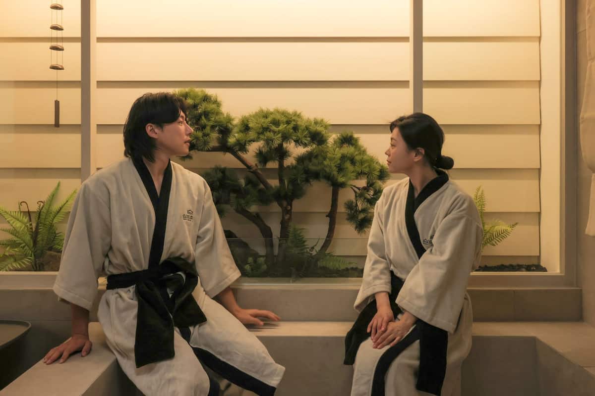 Duas pessoas vestidas com roupas tradicionais de artes marciais sentadas em um interior moderno com design minimalista, uma árvore bonsai e uma luminária na parede.