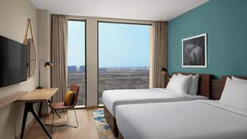 Quarto de hotel com duas camas, uma escrivaninha, uma cadeira, televisão e uma janela grande com vista para a cidade.