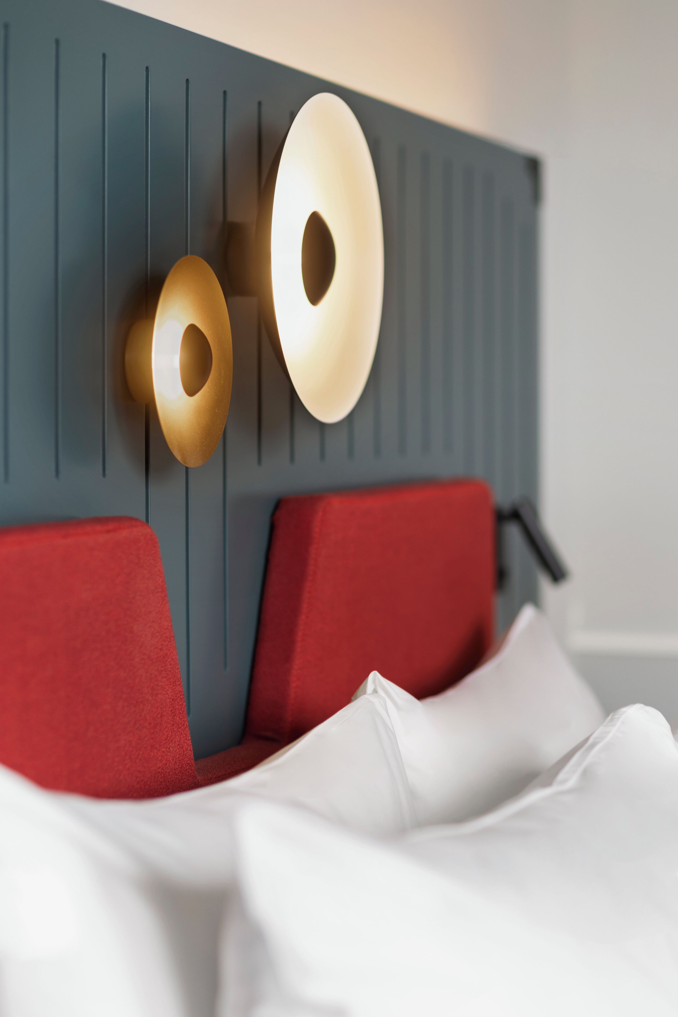 Cama com roupa de cama vermelha e branca, cabeceira cinza com linhas verticais e duas luminárias fixadas na parede.