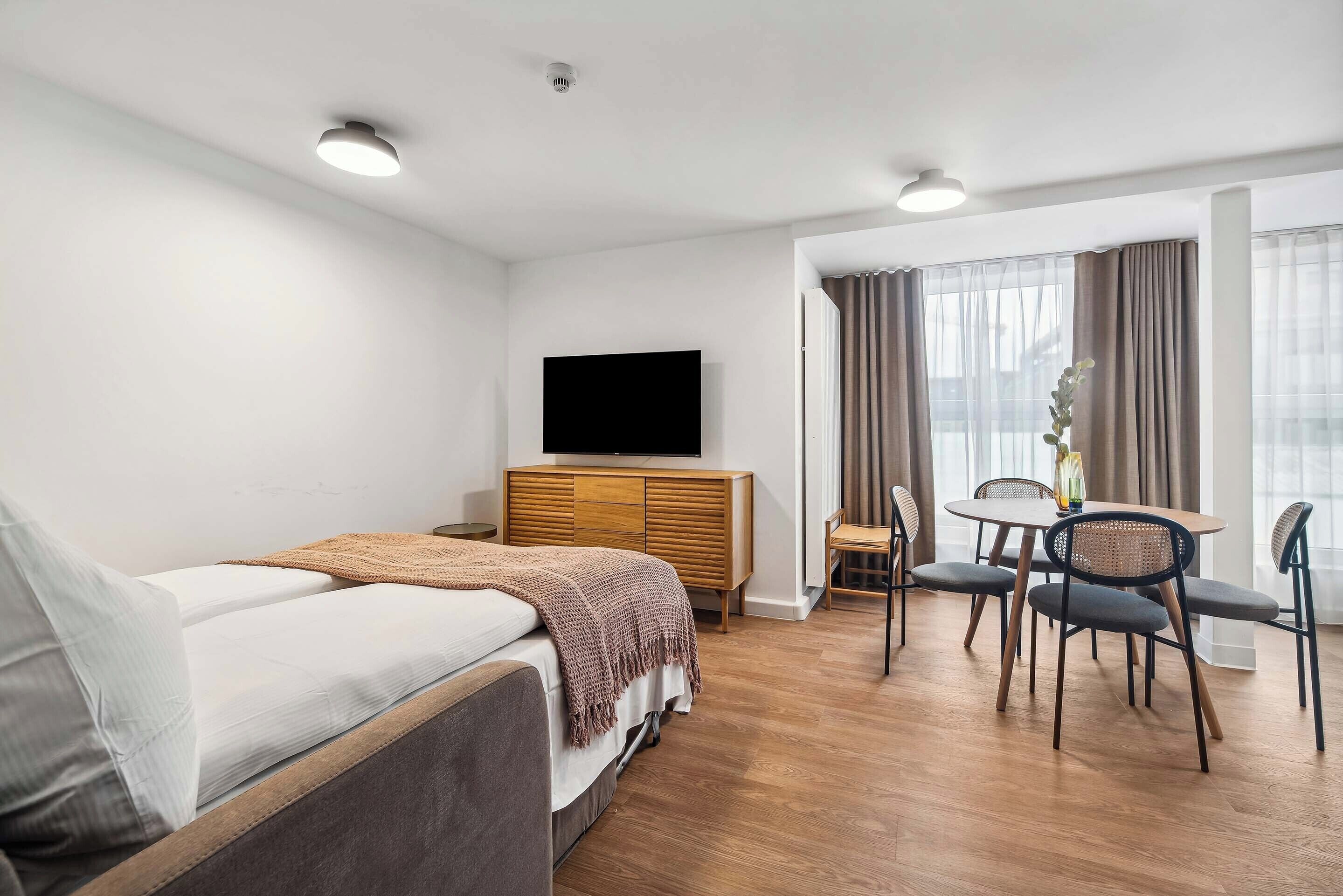 Quarto de hotel com cama, TV fixada na parede, cômoda de madeira, mesa de jantar com cadeiras e uma janela grande com cortinas.
