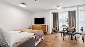 Quarto de hotel com cama, TV fixada na parede, cômoda de madeira, mesa de jantar com cadeiras e uma janela grande com cortinas.