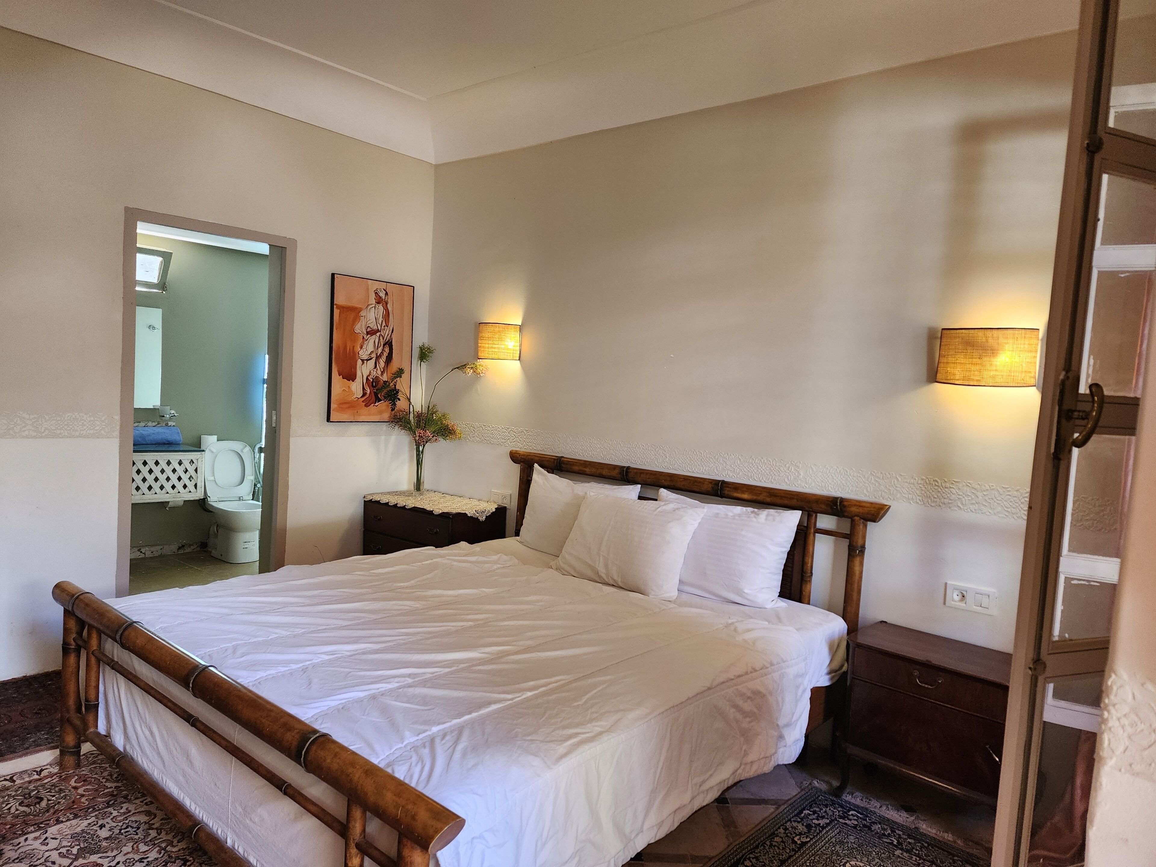 Quarto com cama de madeira, roupa de cama branca, mesa de cabeceira, luminária na parede e uma pintura.