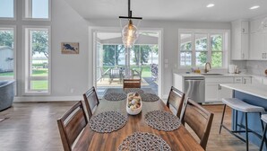 Uma cozinha moderna com armários brancos, uma área de refeições com mesa e cadeiras de madeira, e vista para o exterior através de amplas janelas.