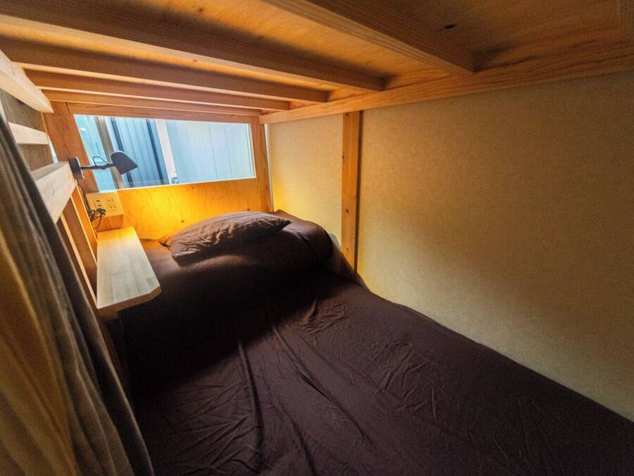 Quarto pequeno e compacto com uma cama de solteiro, mesa de cabeceira e janela que oferece iluminação natural.