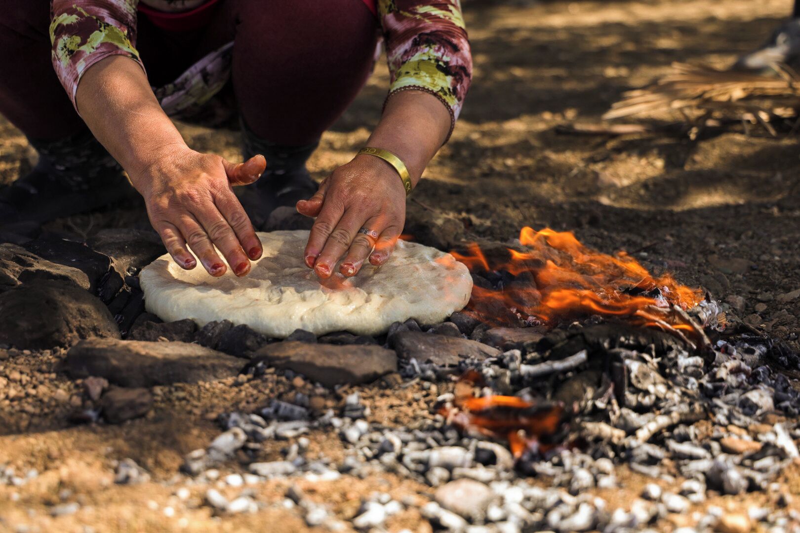 Uma pessoa ajoelhada perto de uma fogueira, preparando comida sobre as chamas.