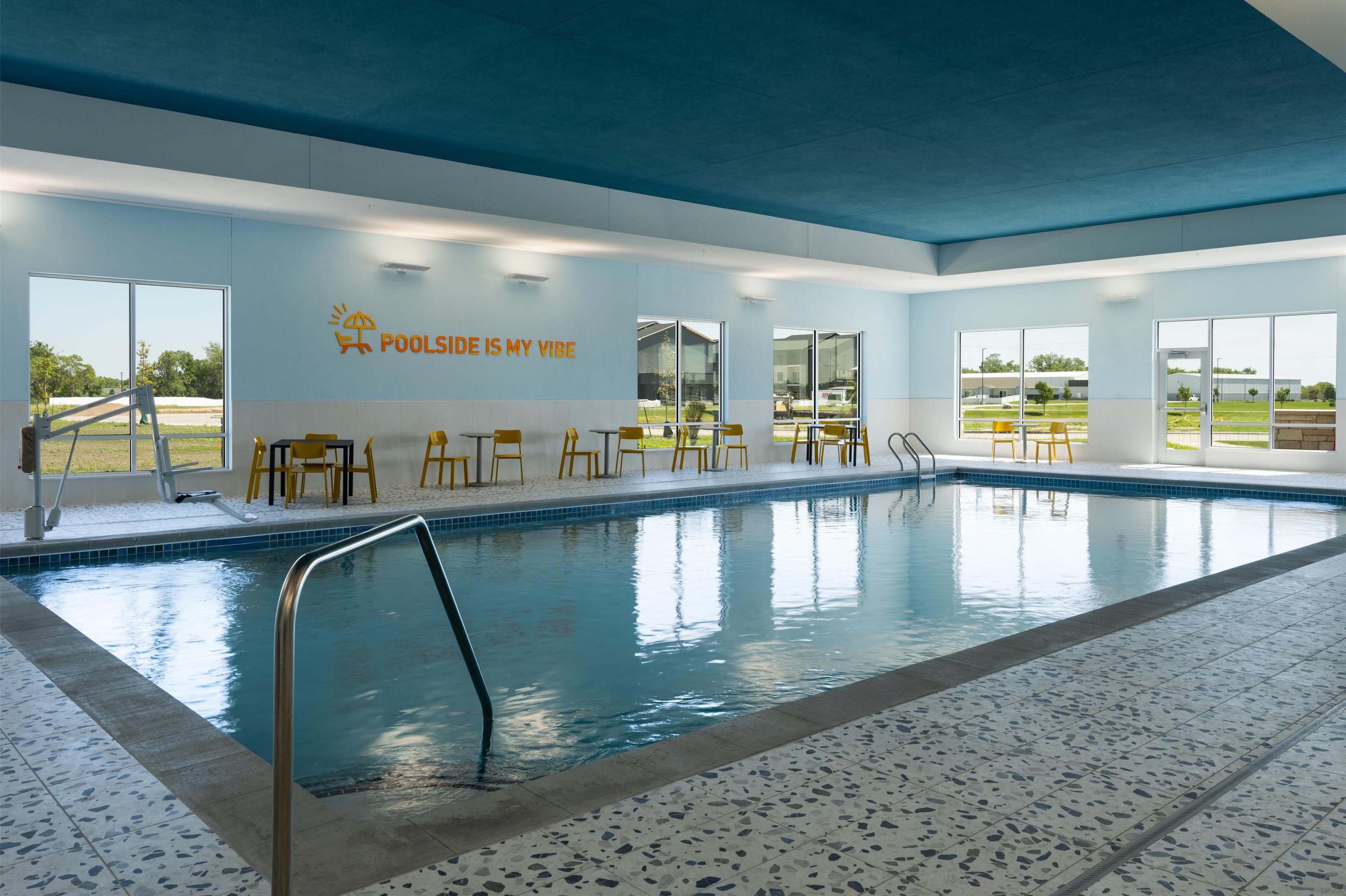 Uma piscina interior com vista desobstruída para o exterior, um logotipo na parede, e um conjunto de cadeiras e mesas amarelas.