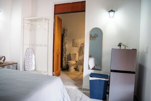 Quarto com cama, guarda-roupa, espelho, pia e sanita visível através de uma porta aberta.