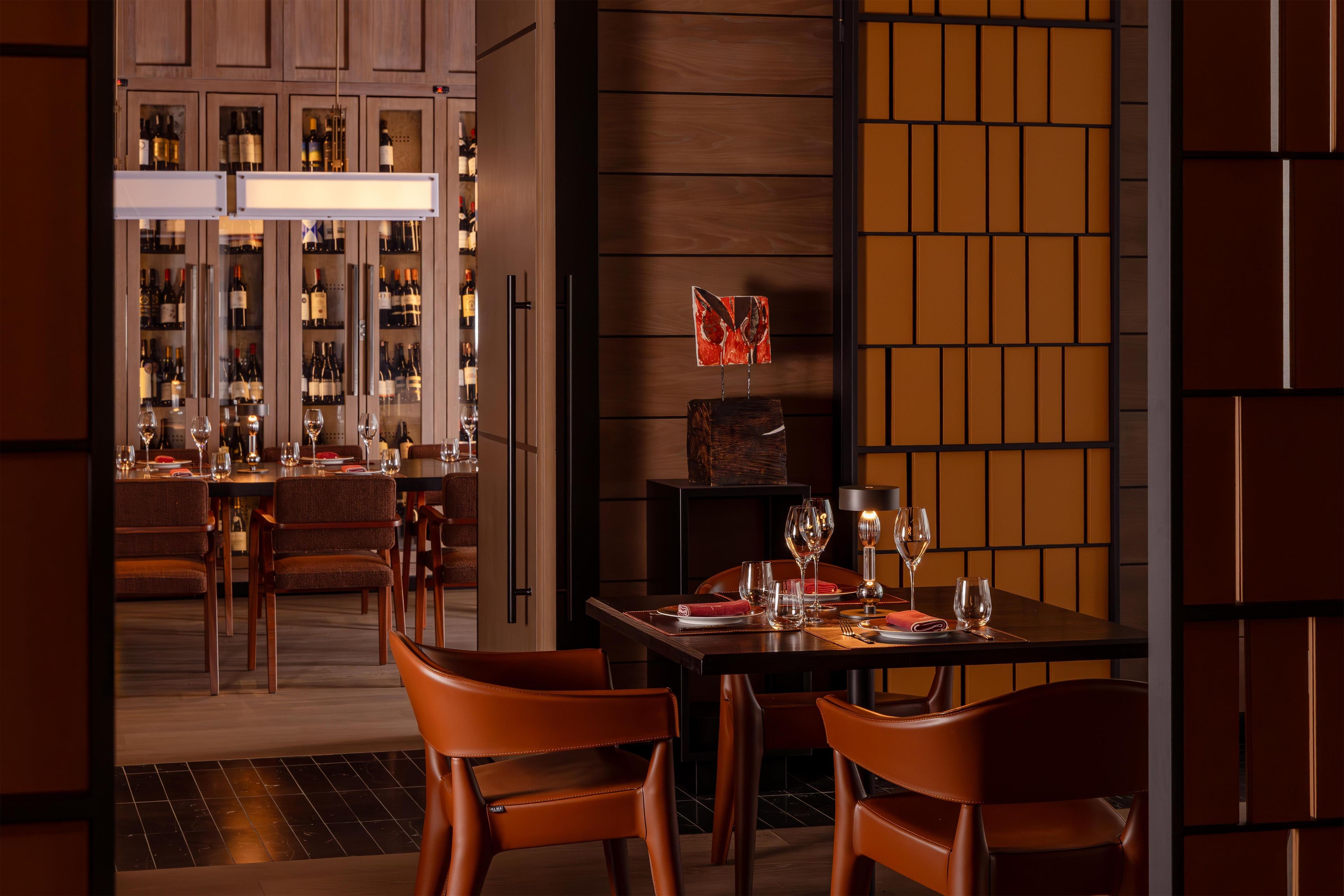 Interior de restaurante com área de bar visível através de uma divisória de vidro, com garrafas e copos de vinho.