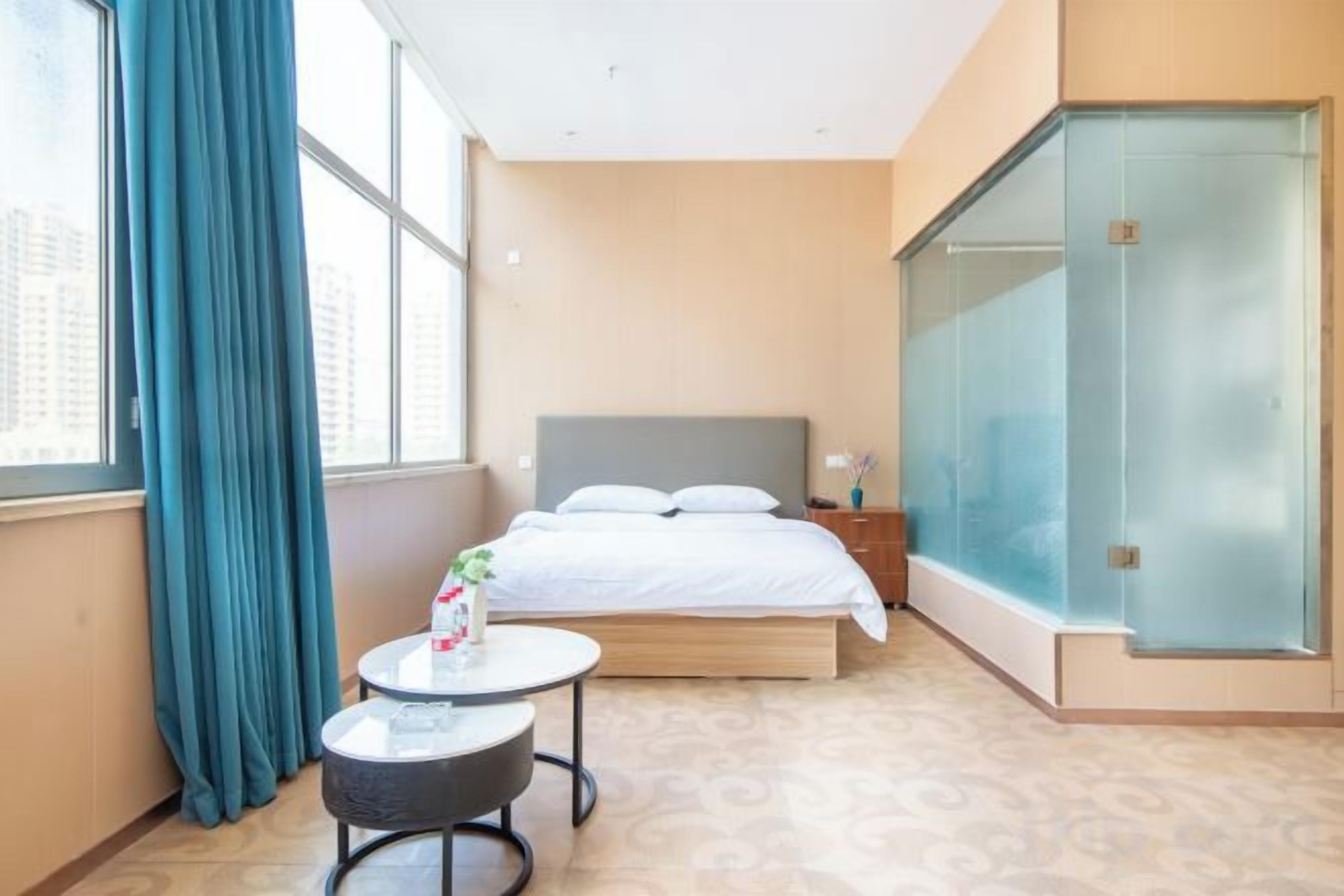 Quarto de hotel com cama, mesa redonda pequena, mesa de cabeceira, janela com cortinas e porta de vidro.