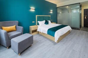 Quarto de hotel moderno com cama, poltrona cinza, mesinha e divisória de vidro.