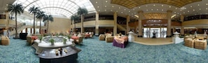 Um amplo hall de hotel com teto alto, vários níveis e área de estar central com mesas e cadeiras.