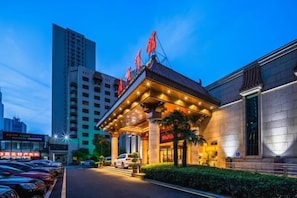 Entrada de hotel com estilo arquitetônico tradicional, estacionamento e um prédio alto ao fundo.