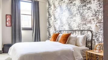 Uma cama bem arrumada com almofadas brancas e laranja, uma cabeceira de metal preto e uma parede com papel de parede estampado com flores.