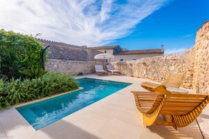 Piscina com espreguiçadeiras e uma mesa, cercada por muros de pedra e vegetação.