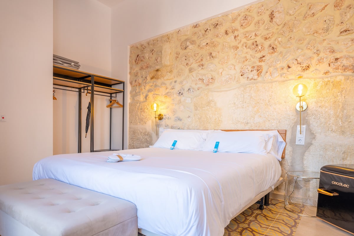 Quarto com parede de pedra, cama com roupa de cama branca, um banco, uma mala e um cabideiro.