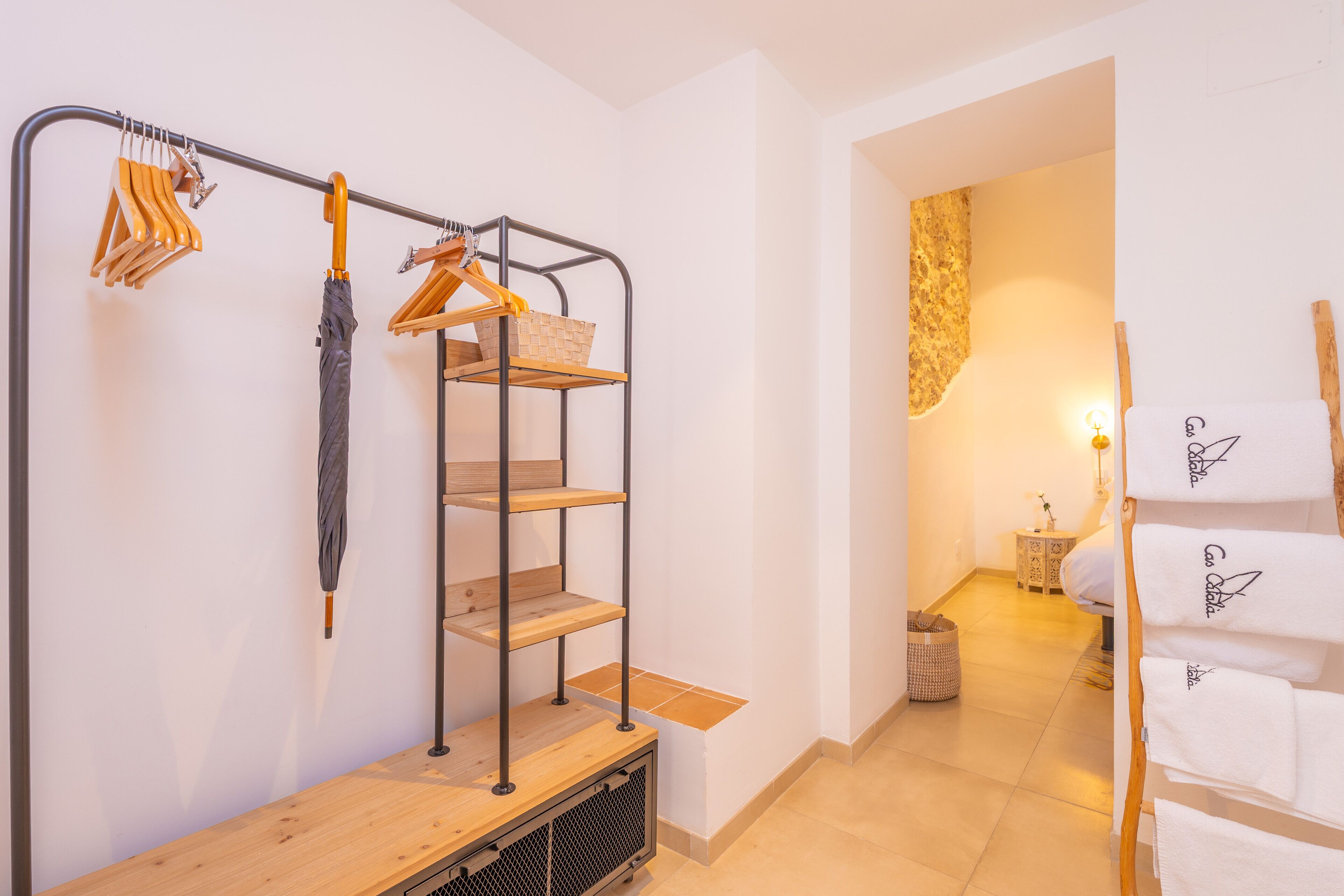 Quarto compacto com uma estante metálica contendo cabides de madeira e um guarda-chuva cinzento, uma prateleira de madeira com um cesto, e uma pequena mesa com um cesto. Há um porta-toalhas fixado na parede com toalhas brancas.