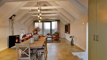 Um amplo loft com teto alto, vigas aparentes, área de jantar com uma mesa comprida e cadeiras, lareira e uma grande janela com vista.