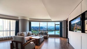 Uma sala de estar espaçosa com uma grande janela que oferece vista para a cidade, um conjunto de sofás e uma televisão de tela plana fixada na parede.
