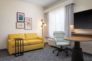 Quarto de hotel com um sofá amarelo, uma poltrona estampada, uma mesa pequena e obras de arte emolduradas na parede.