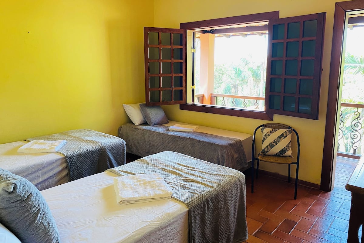 Quarto com duas camas de solteiro, uma cadeira, janela com persianas e vista para o verde lá fora.