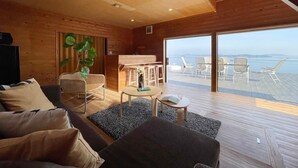 Um interior moderno com teto de madeira, área de estar com sofá e cadeiras, uma pequena mesa com um vaso, e vista para o mar por meio de grandes janelas.