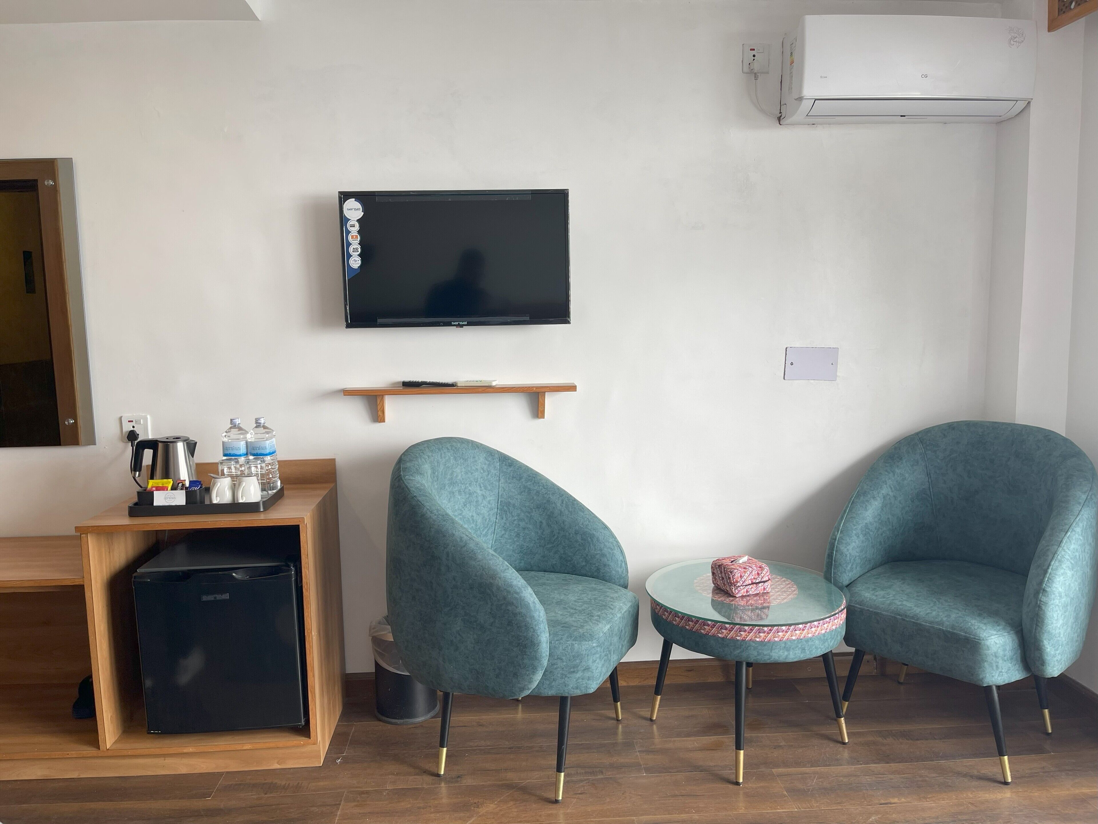 Uma sala de estar moderna com televisão de tela plana, dois poltronas verde-água, uma mesa redonda pequena, uma máquina de café e um mini-refrigerador.