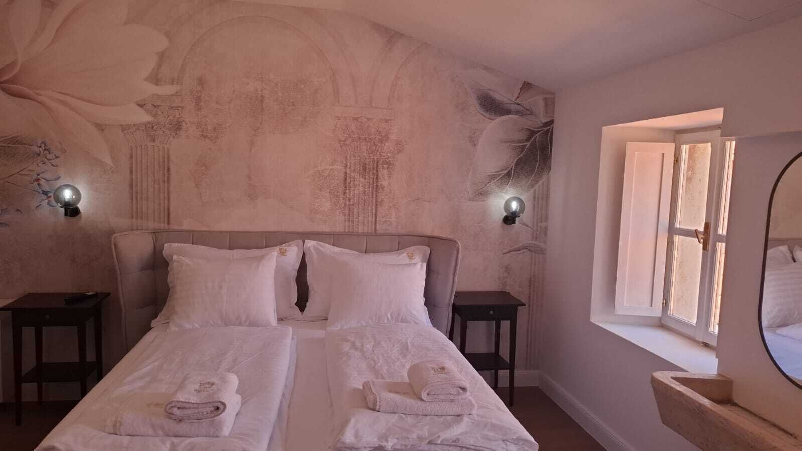 Uma cama de casal com roupa de cama e toalhas brancas, duas mesas de cabeceira, um aplique na parede e uma janela com vista para o exterior.