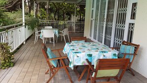 Um alpendre coberto com mesa e cadeiras, cercado por um corrimão branco e vegetação.