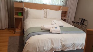 Quarto de hotel com uma cama grande, mesa de cabeceira, cadeira e janela com cortinas.