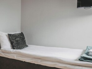 Uma cama de solteiro com colchão branco e travesseiro cinza, um cobertor dobrado e uma televisão fixada na parede.