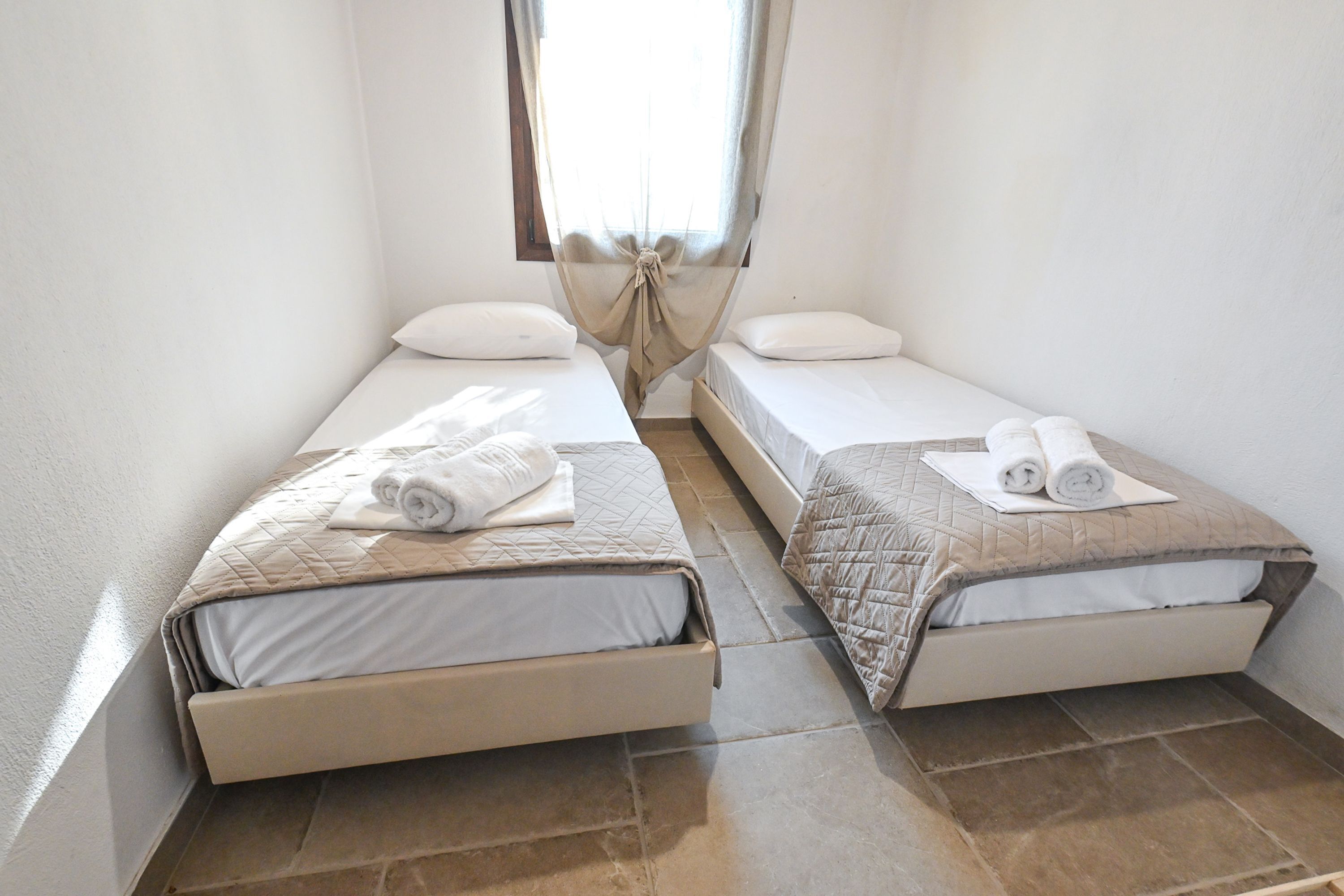 Quarto pequeno e limpo, com duas camas de solteiro, uma janela com cortinas e piso ladrilhado.