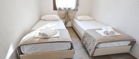 Quarto pequeno e limpo, com duas camas de solteiro, uma janela com cortinas e piso ladrilhado.